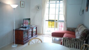 Uma sala de estar com uma televisão em um móvel de madeira, uma mesa branca, um sofá vermelho e uma janela com cortinas.