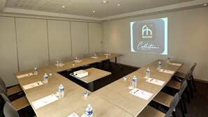 Uma sala de conferências com uma grande mesa, cadeiras, garrafas de água e uma tela de projetor exibindo o logotipo "fasthotel Collection".