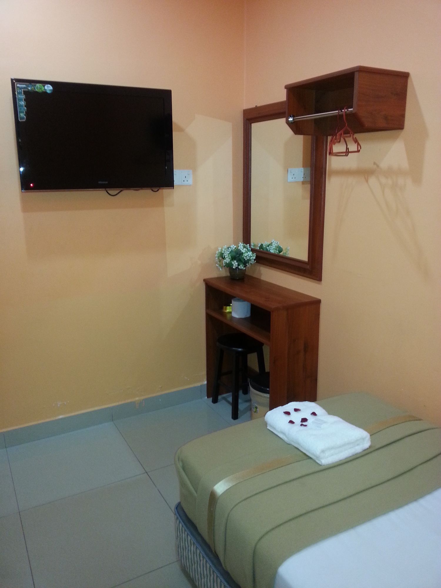 Quarto de hotel com cama, televisão na parede, prateleira de madeira com espelho e um banquinho pequeno.
