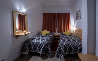 Quarto com duas camas, cada uma com roupa de cama estampada e um travesseiro amarelo. Há um espelho na parede, uma pequena prateleira com uma vela e um quadro emoldurado.