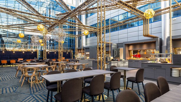Uma sala de jantar moderna com várias mesas e cadeiras, uma cozinha ao fundo e um teto alto com vigas aparentes e luminárias pendentes.