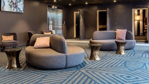 Um lobby moderno com carpete estampado em azul, sofás cinzas com almofadas e uma tela grande exibindo uma cena de filme.