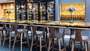 Uma área de bar com várias cadeiras e mesas, um mostrador de vinhos ao fundo e um tapete estampado.