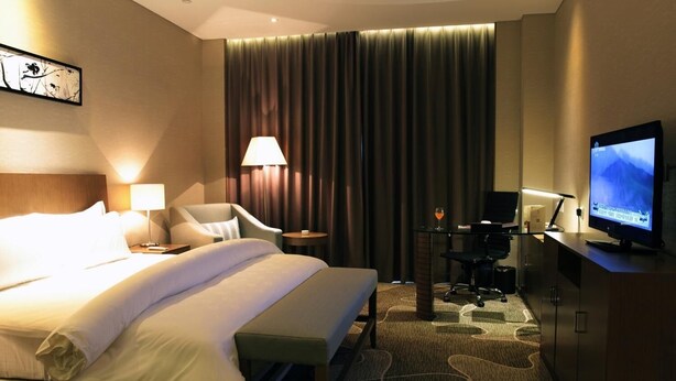 Quarto de hotel com cama grande, escrivaninha, cadeira, televisão e janela com cortinas.