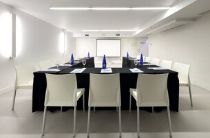 Sala de conferência com uma mesa comprida, cadeiras brancas, toalhas de mesa pretas, garrafas de água azuis e uma tela de projetor.