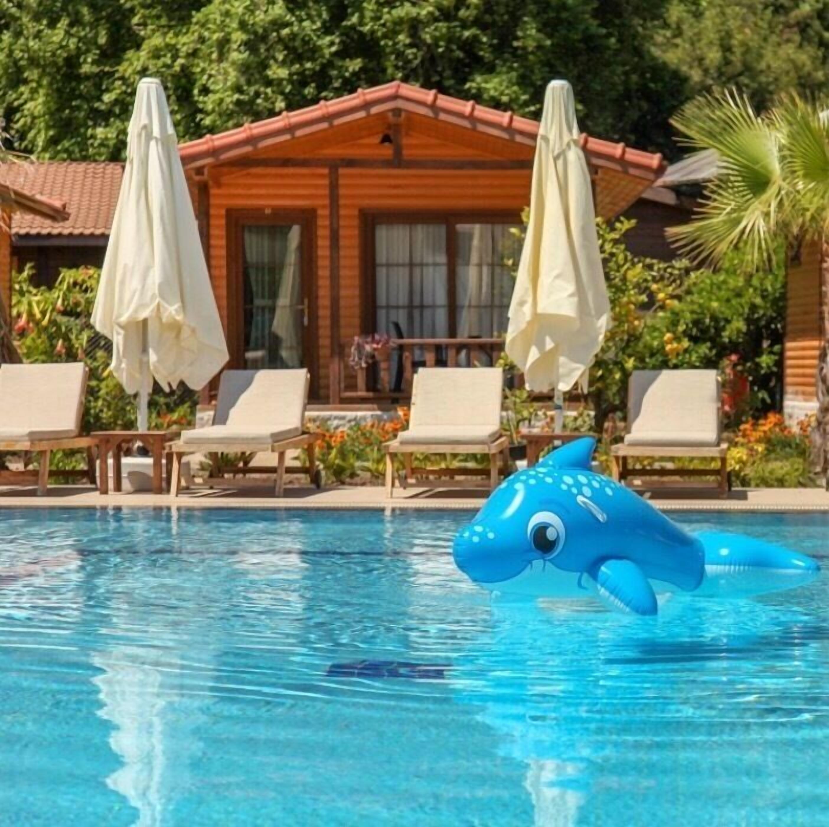 Uma piscina com espreguiçadeiras e guarda-sóis, um prédio de madeira com janelas e uma palmeira ao fundo.