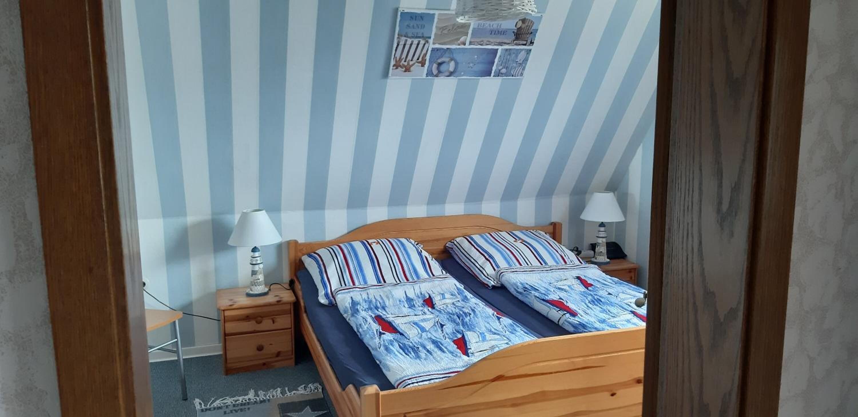 Quarto com parede listrada, cama de madeira com roupa de cama listrada, duas mesas de cabeceira com abajures e uma parede com várias imagens.