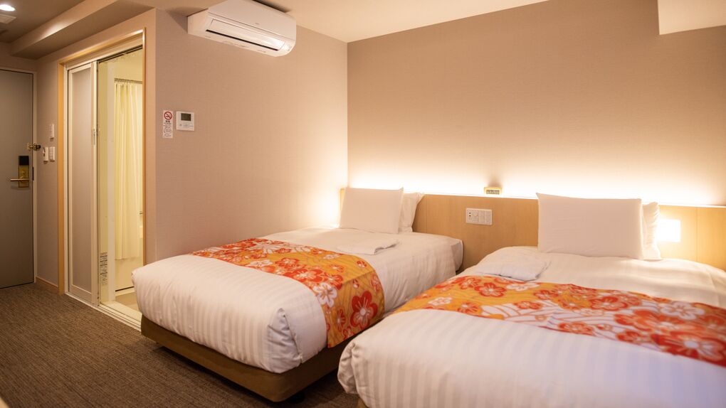 Quarto de hotel com duas camas, ar condicionado, uma porta e luminária fixada na parede.