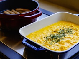 Uma travessa retangular com um prato amarelo e cremoso, possivelmente um gratinado, decorada com ervas, sobre uma bancada escura.
