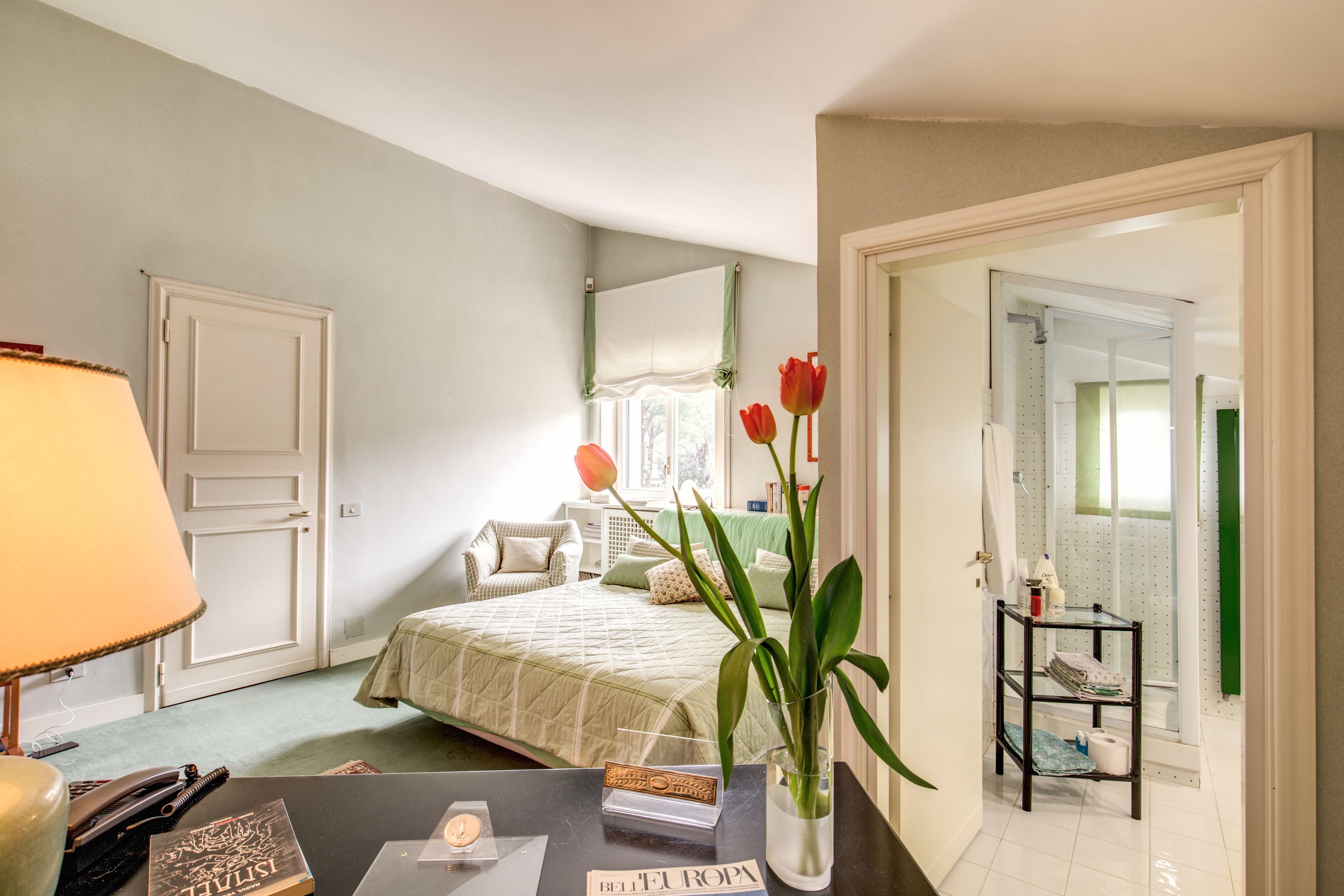 Quarto de hotel com uma cama, uma escrivaninha com abajur, um vaso com flores e um banheiro visível através de uma porta aberta.