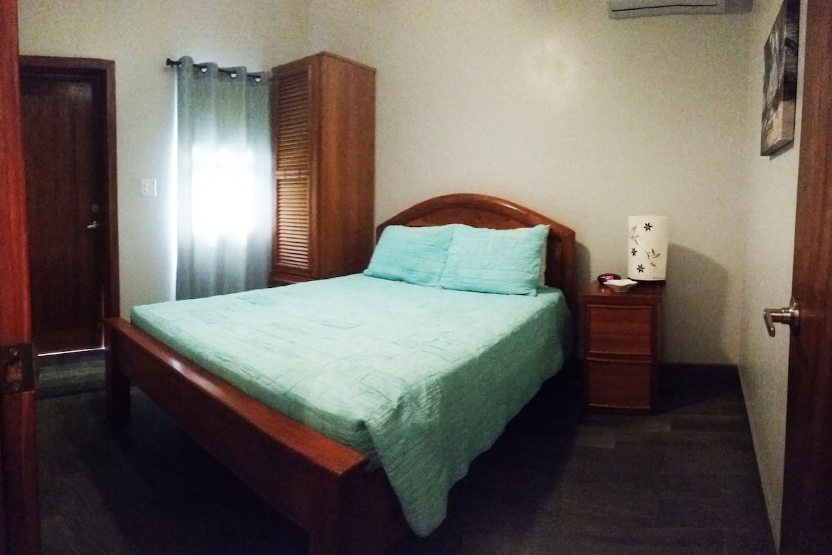 Quarto individual com uma cama de solteiro, cabeceira de madeira, mesa de cabeceira com abajur e janela com cortinas.
