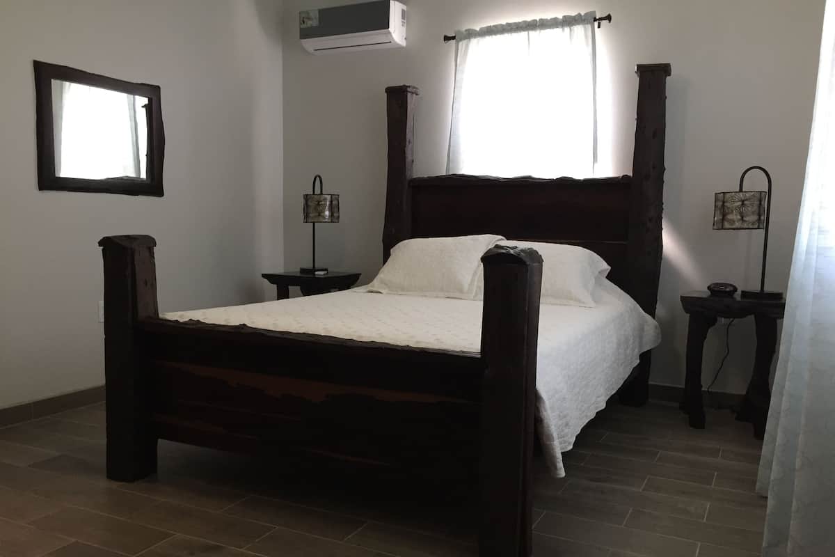 Quarto com cama de madeira, uma mesinha com luminária, ar condicionado na parede e janela com cortinas leves.