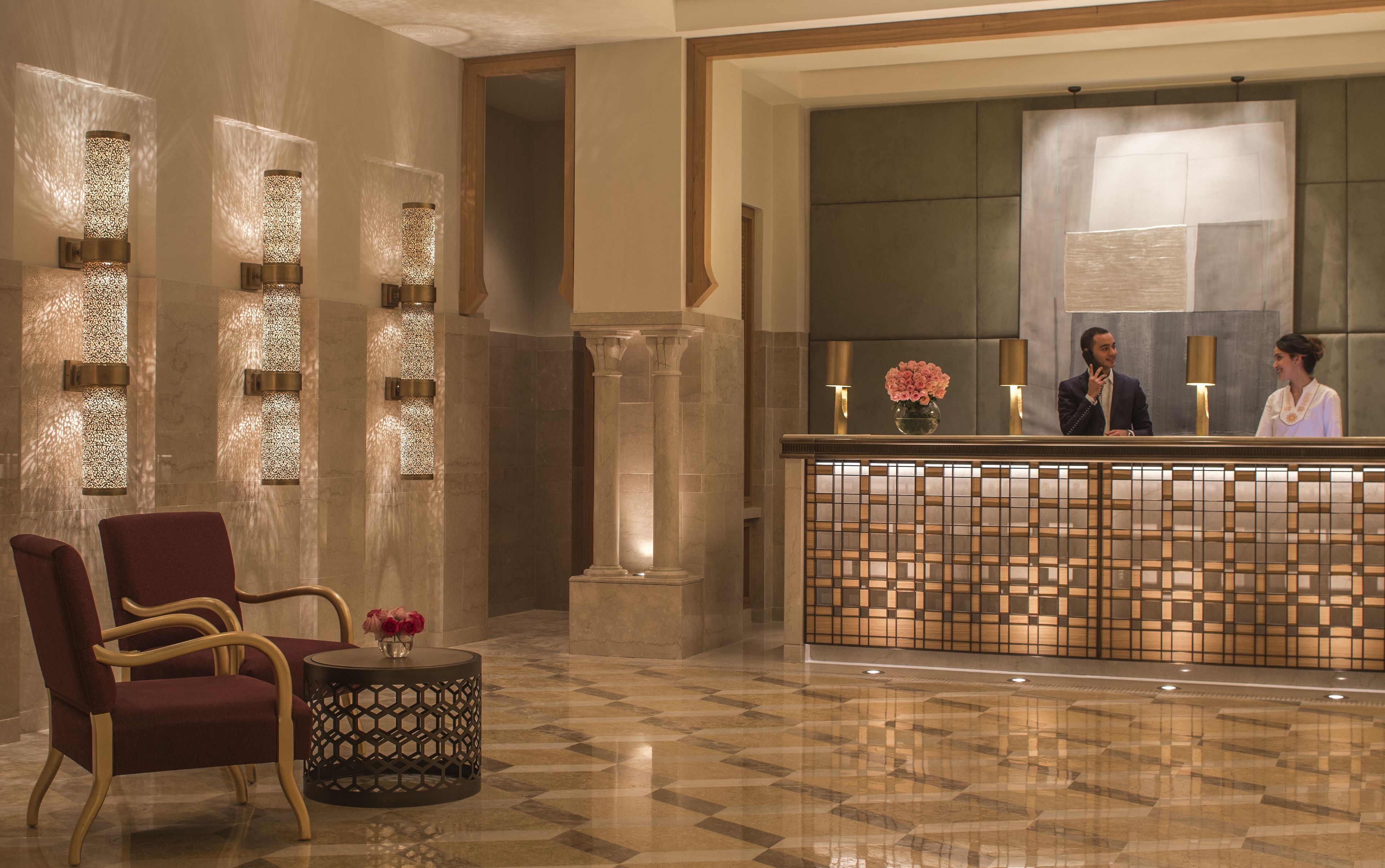 Uma área de recepção com balcão de check-in, espaço de espera com uma cadeira e mesa, e arandelas decorativas na parede.