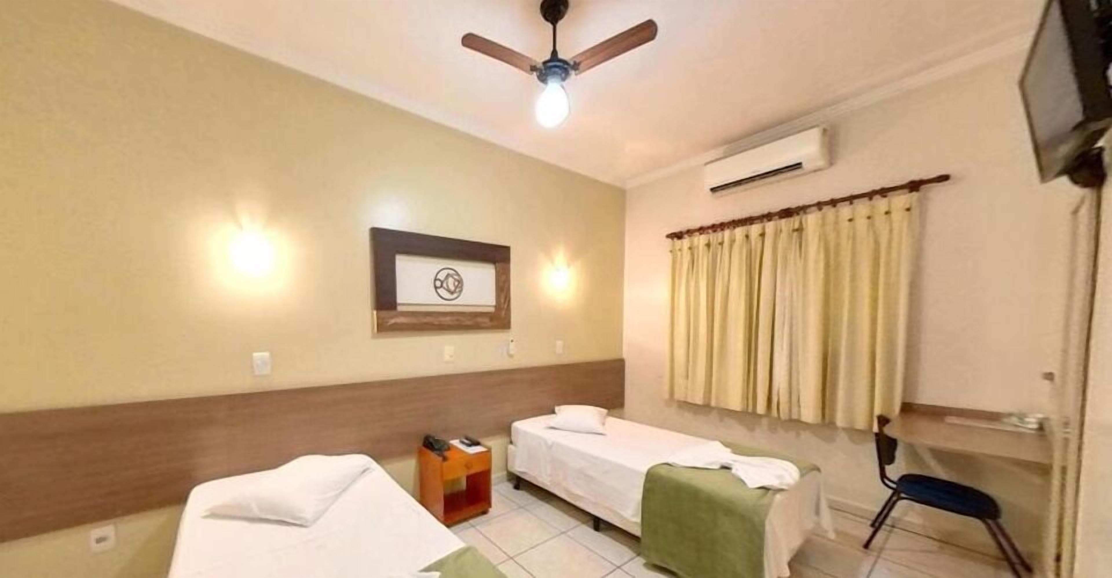 Quarto de hotel com duas camas de solteiro, ventoinha no teto, ar condicionado, quadro emoldurado na parede e janela com cortinas.