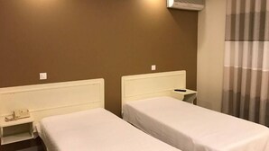 Quarto de hotel com duas camas de solteiro, uma mesa de cabeceira com luminária, ar condicionado e uma janela com cortinas leves.