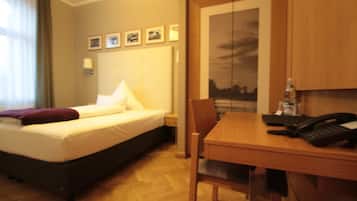 Quarto de hotel com cama, escrivaninha e cadeira. Há quadros nas paredes e uma janela com cortinas.