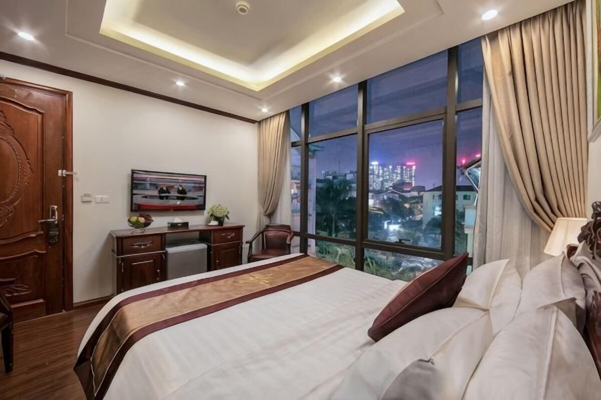 Quarto de hotel com uma cama grande, uma mesa com cadeira, televisão e vista para a paisagem urbana por grandes janelas.