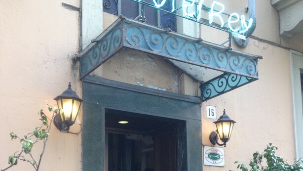 Entrada de um hotel com um letreiro de neon que diz "Hotel Rey" acima da porta. Há duas luminárias na parede, uma de cada lado da entrada.