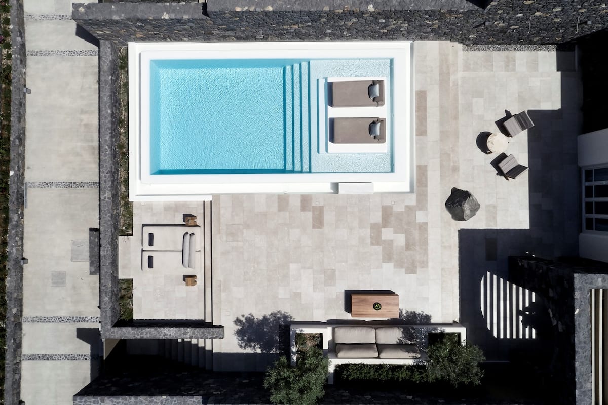 Vista aérea de uma área de piscina moderna, com piscina retangular, duas espreguiçadeiras, um espaço de estar pequeno e uma parede de pedra.