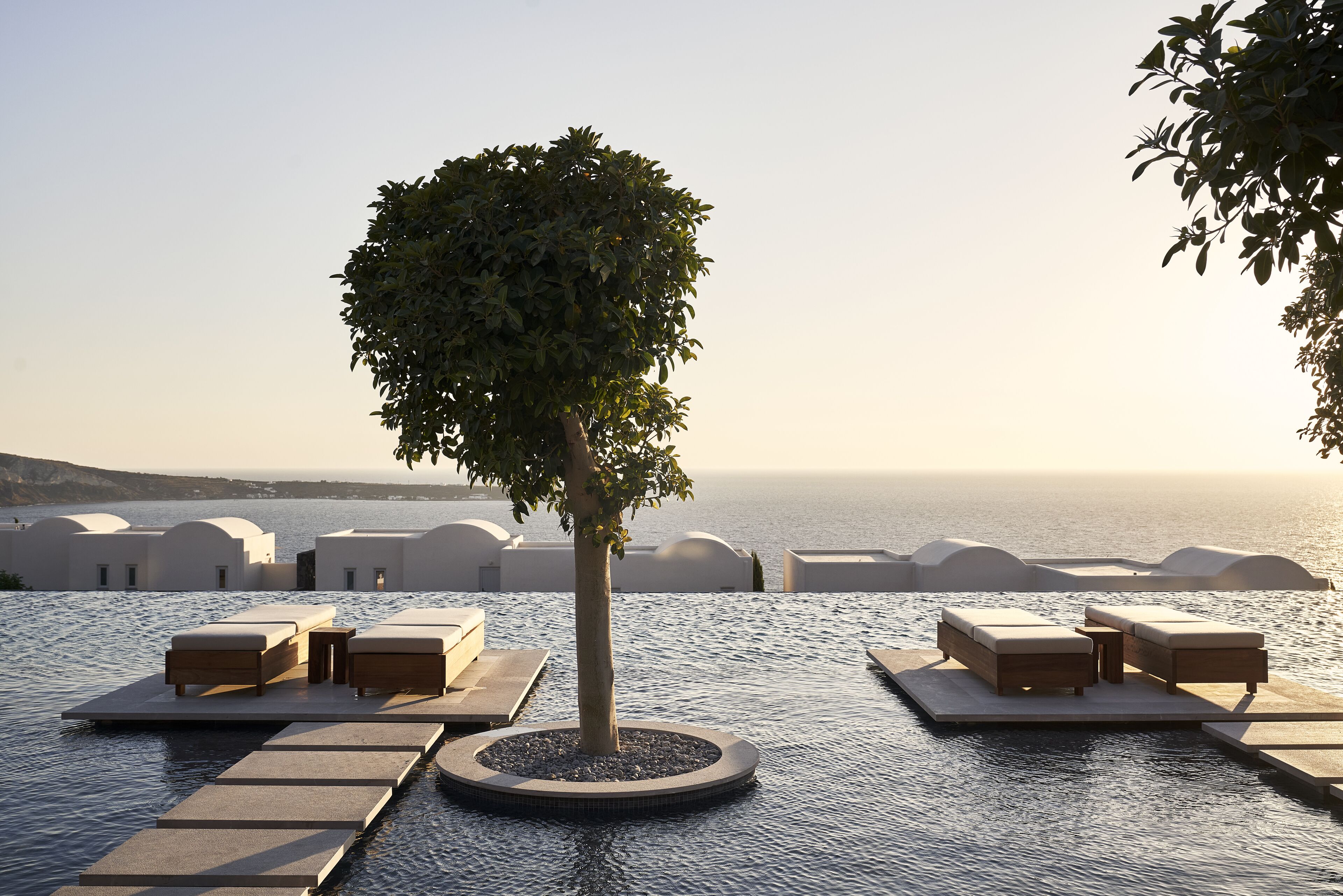 Área à beira da piscina com espreguiçadeiras e uma árvore, com vista para um corpo d'água e edifícios ao fundo.