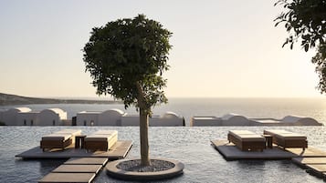Área à beira da piscina com espreguiçadeiras e uma árvore, com vista para um corpo d'água e edifícios ao fundo.