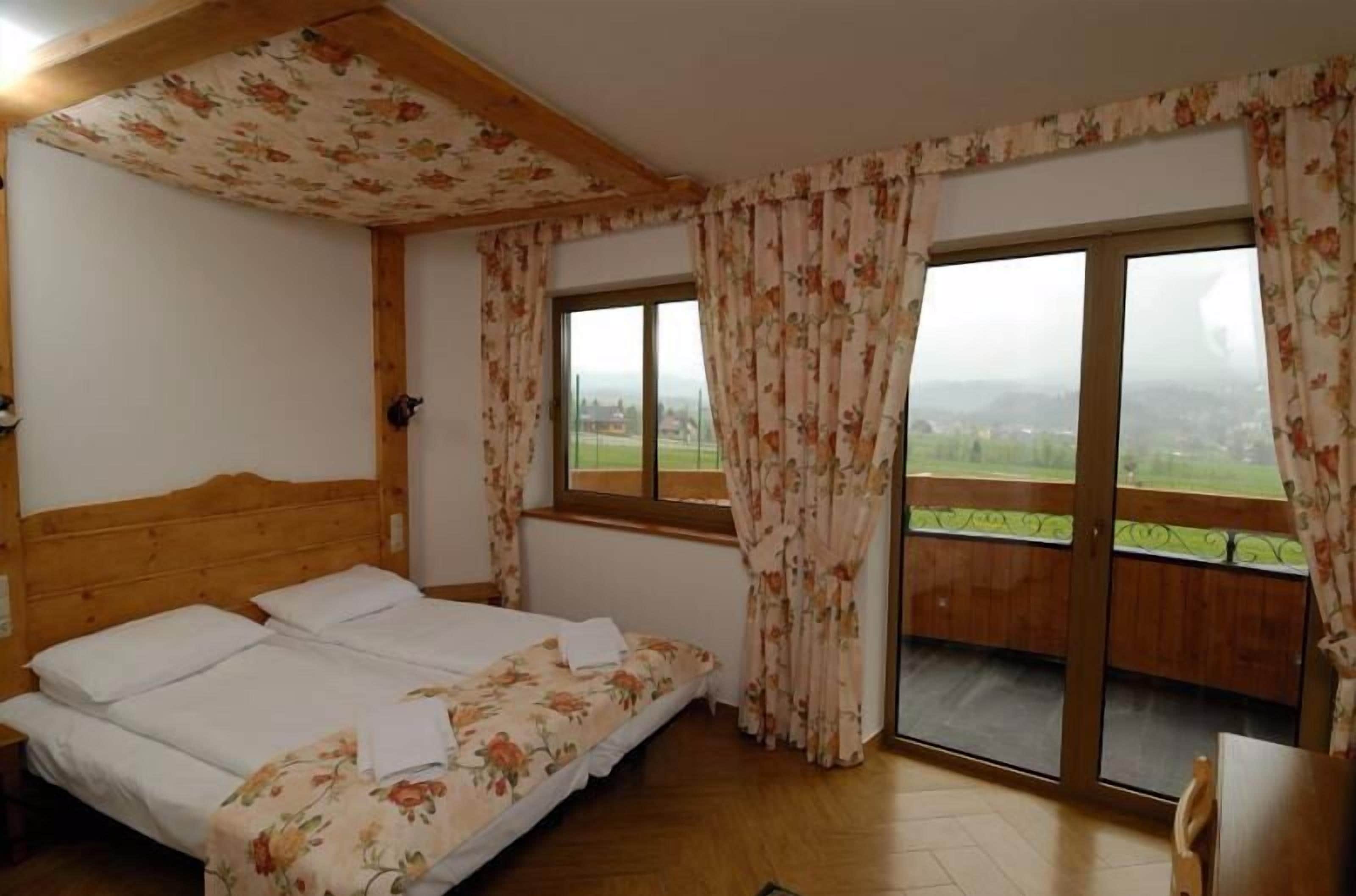 Quarto com cabeceira de madeira, cama com roupa de cama floral, janela grande com cortinas e vista para uma paisagem verde.