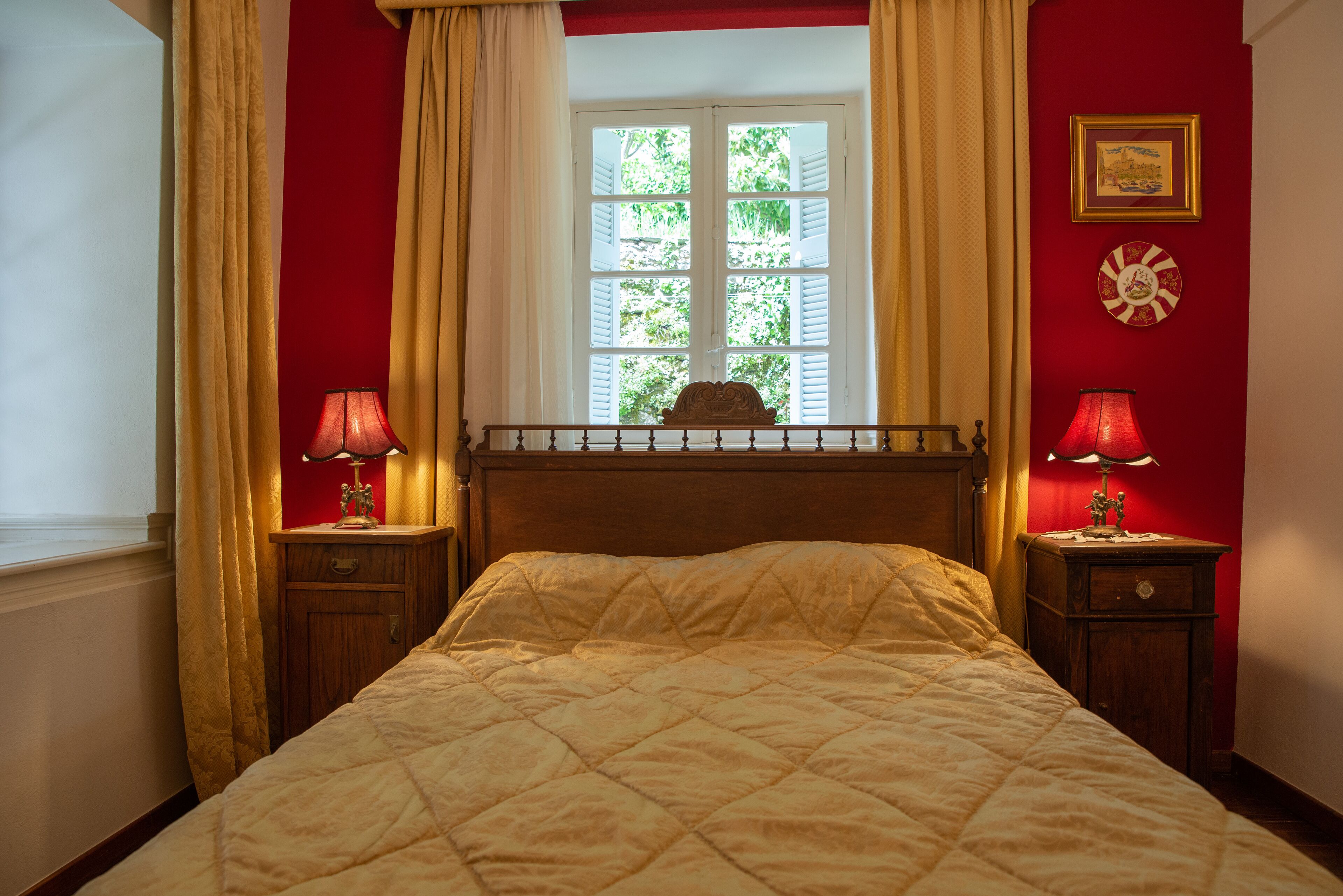 Quarto com cama de madeira, duas mesas de cabeceira com luminárias, janela com cortinas e parede vermelha com um quadro emoldurado.