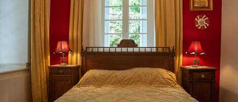 Quarto com cama de madeira, duas mesas de cabeceira com luminárias, janela com cortinas e parede vermelha com um quadro emoldurado.