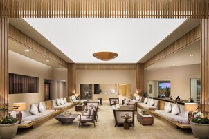 Um hall de hotel espaçoso, com teto alto, painéis de madeira e um lustre central. Há várias áreas de estar com sofás e poltronas, e um balcão de recepção ao fundo.