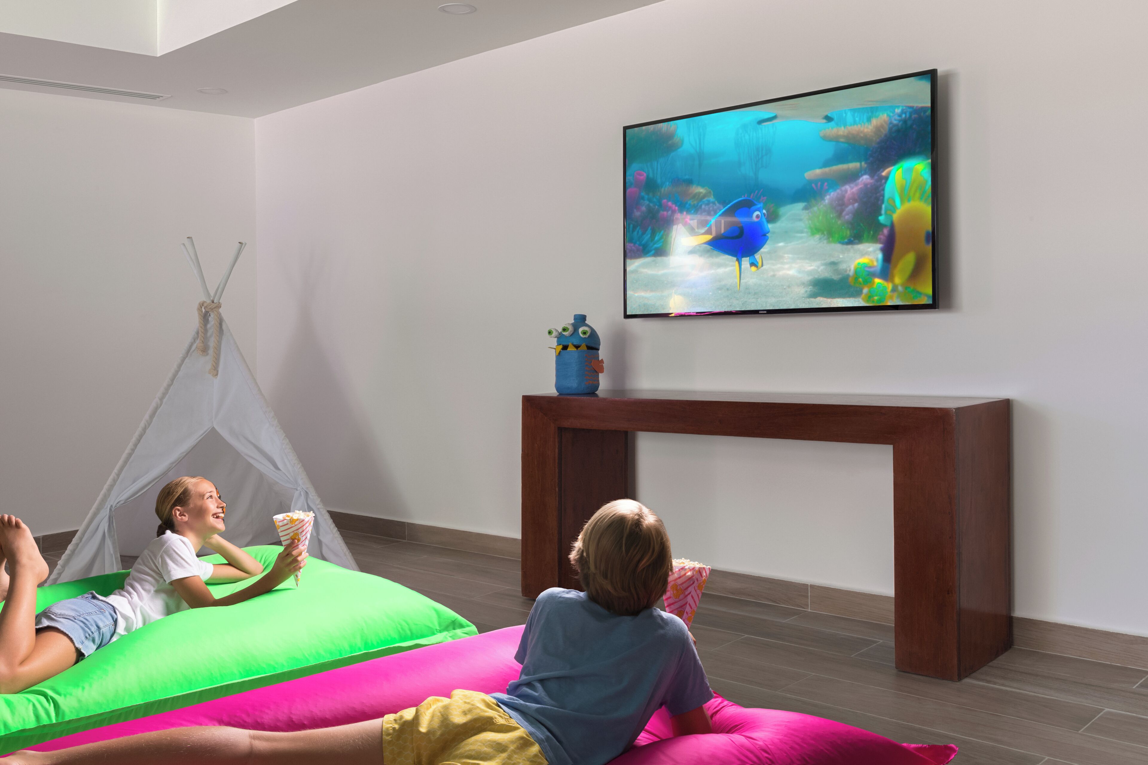 Um quarto com televisão em suporte, uma cama, um sofá verde e rosa, e uma pequena mesa com um objeto azul.
