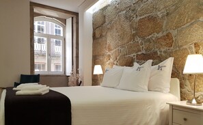 Uma cama bem arrumada com lençóis e almofadas brancos, um cobertor preto e uma janela com vista para edifícios. Há duas luminárias de cabeceira em uma mesa de cabeceira.