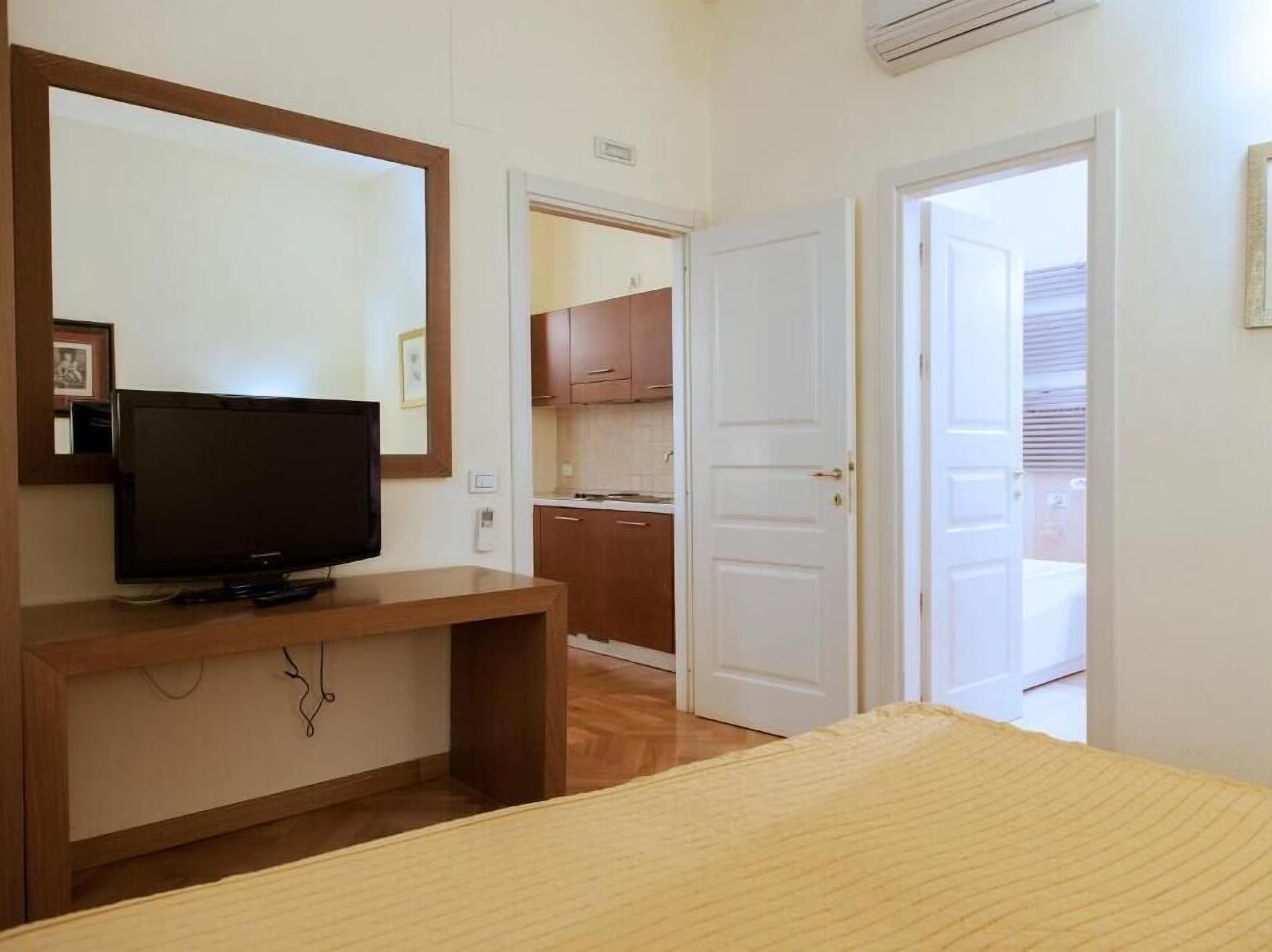 Quarto com uma cama, uma televisão sobre um suporte de madeira, um espelho e uma porta que dá para a cozinha.