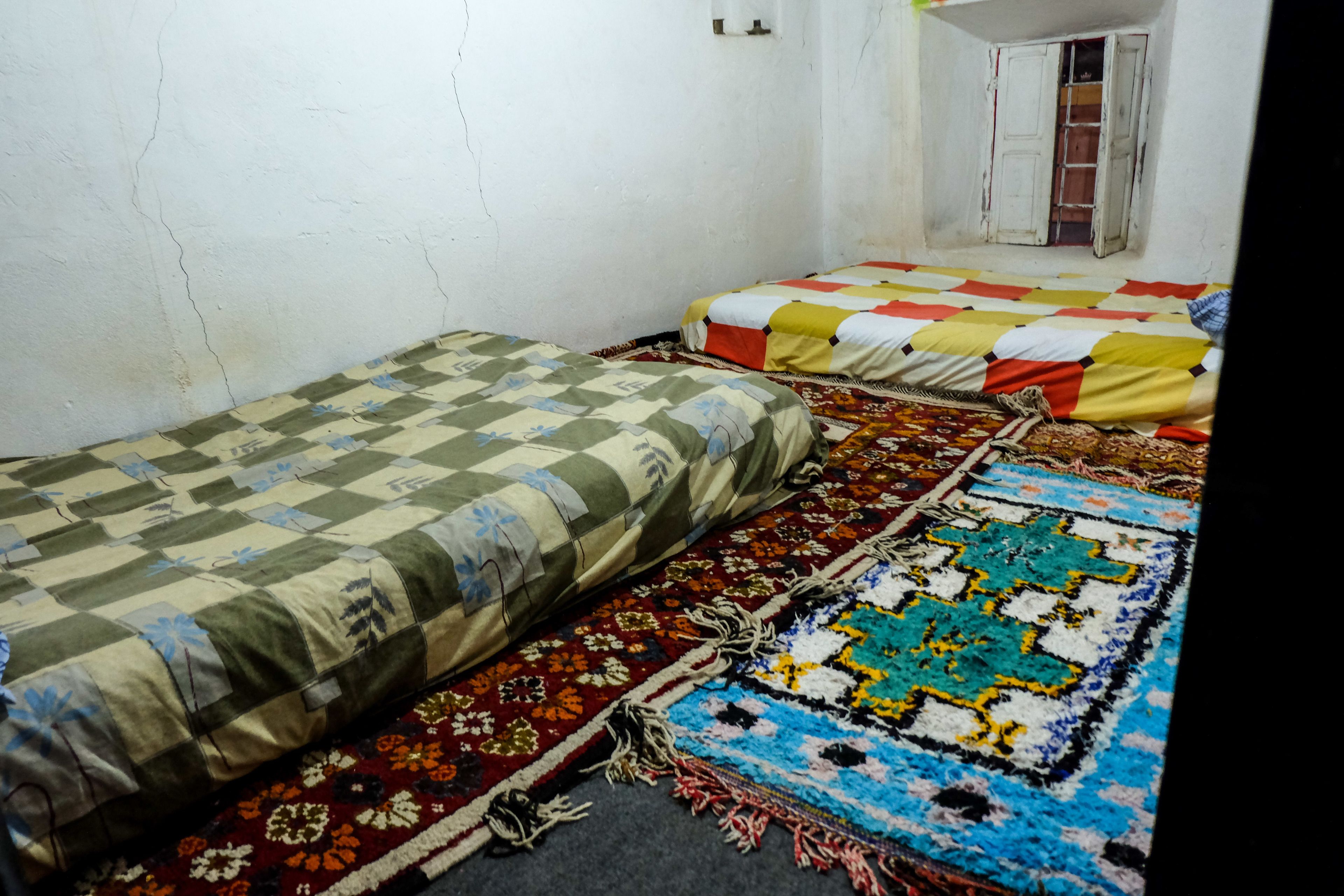 Quarto com duas camas, cada uma com um edredom de padrão diferente. Há uma pequena janela com uma persiana branca e as paredes são lisas, com rachaduras visíveis.