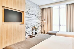Quarto de hotel moderno com uma televisão de tela plana grande fixada em uma parede de madeira, uma parede de destaque com textura de pedra, um vaso com flores sobre uma mesa de cabeceira e uma janela grande com cortinas.