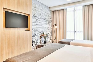 Quarto de hotel moderno com uma televisão de tela plana grande fixada em uma parede de madeira, uma parede de destaque com textura de pedra, um vaso com flores sobre uma mesa de cabeceira e uma janela grande com cortinas.