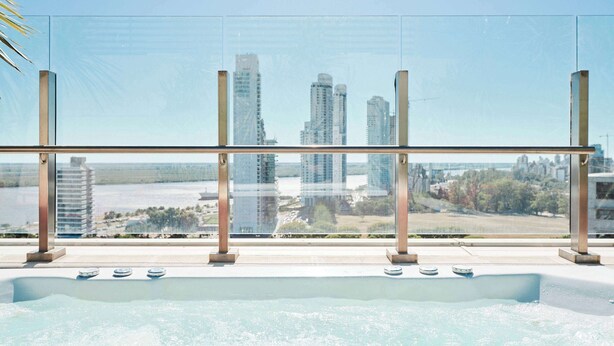 Uma piscina no terraço com guarda-corpo de vidro, oferecendo vista para edifícios altos e um corpo de água.