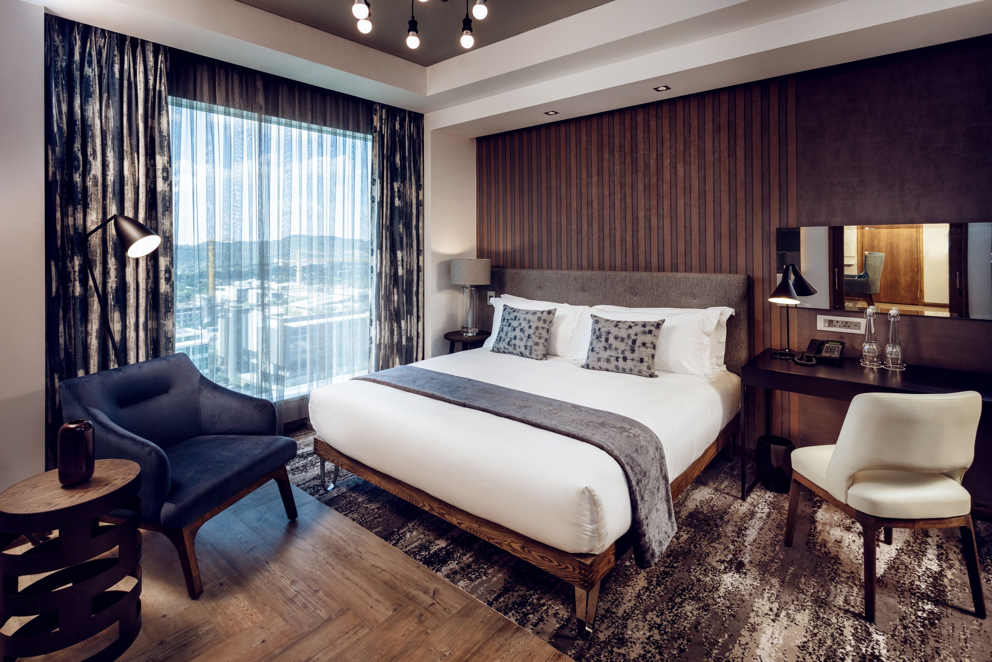 Quarto de hotel moderno com uma cama grande, uma escrivaninha, duas cadeiras, uma luminária e vista para a cidade.