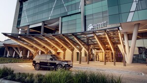 Entrada moderna de hotel com fachada de vidro, área externa coberta com guarda-sóis e um SUV de luxo estacionado em frente.