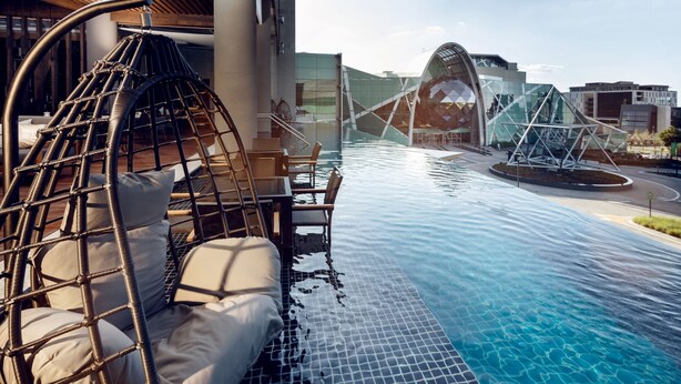 Uma área de piscina moderna em hotel, com uma cadeira suspensa, espreguiçadeiras e um edifício envidraçado ao fundo.