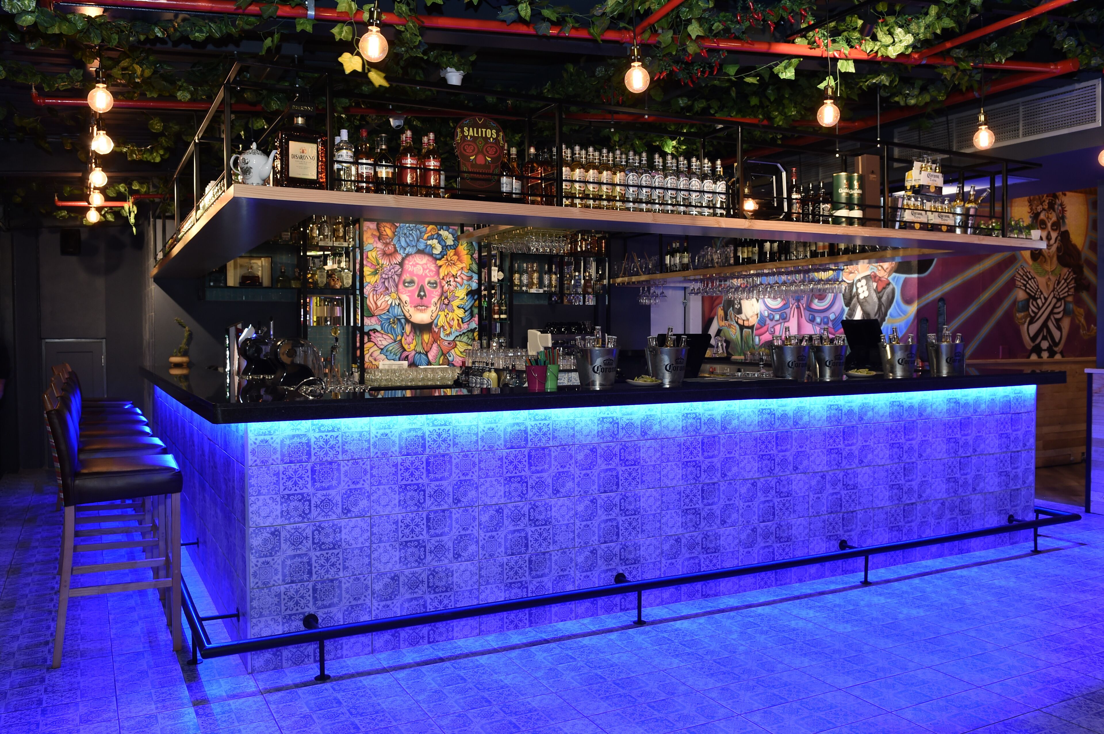 Uma área de bar com balcão revestido de azulejos, prateleiras cheias de garrafas e um mural.