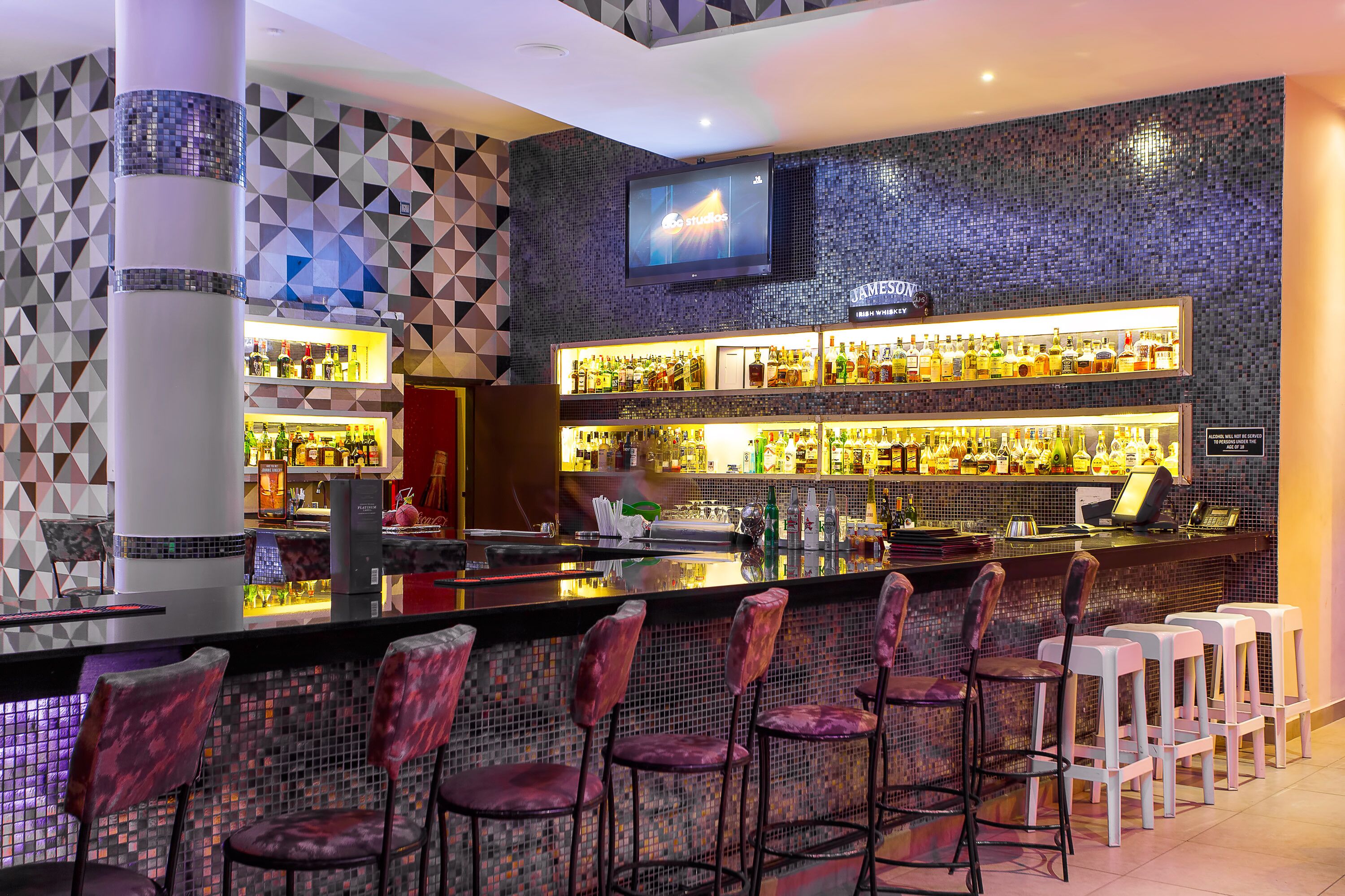 Uma área de bar com design moderno, destacando uma parede com padrão geométrico, uma TV de tela plana e uma variedade de garrafas de bebidas.