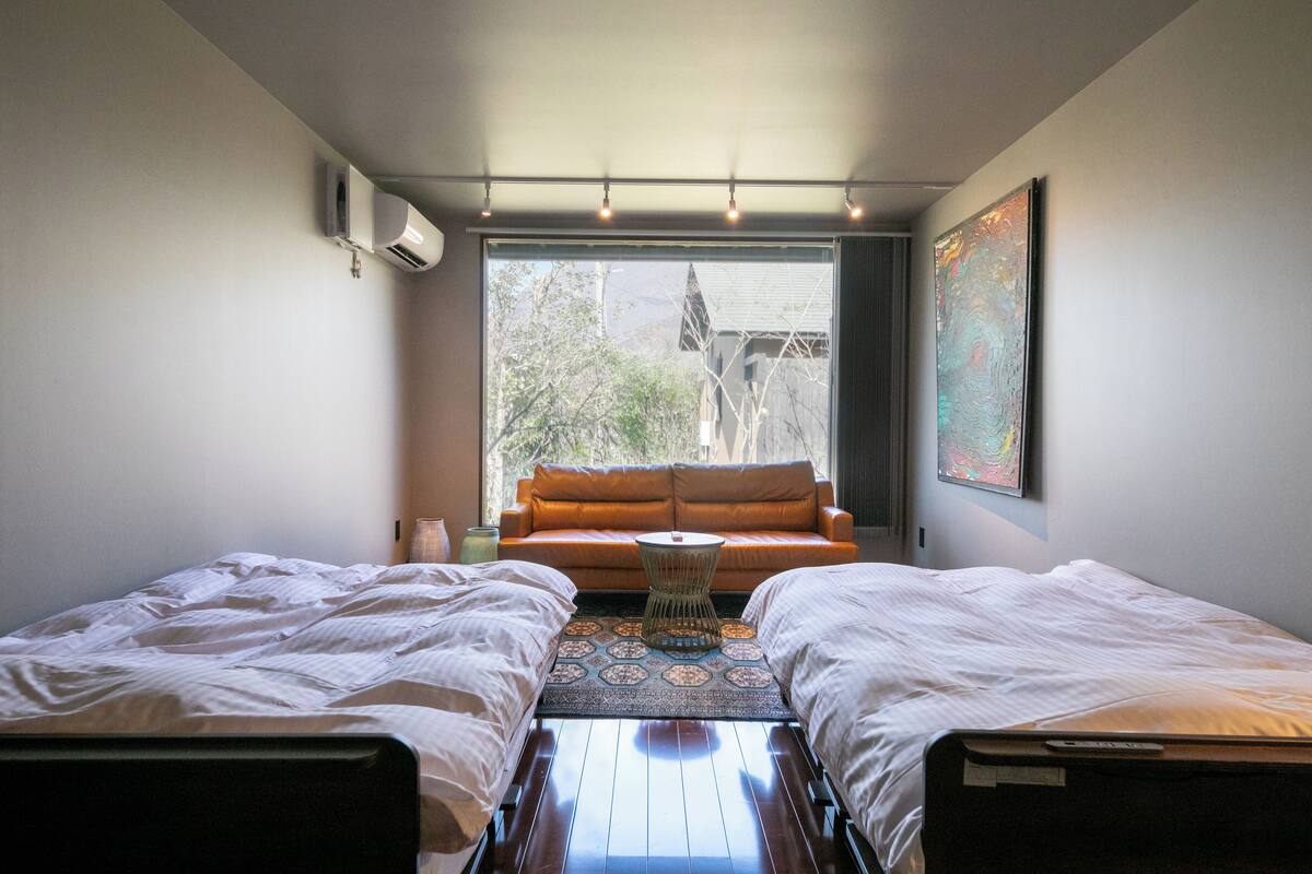 Quarto com duas camas, um sofá, um tapete estampado e uma janela grande com vista para edifícios e árvores.