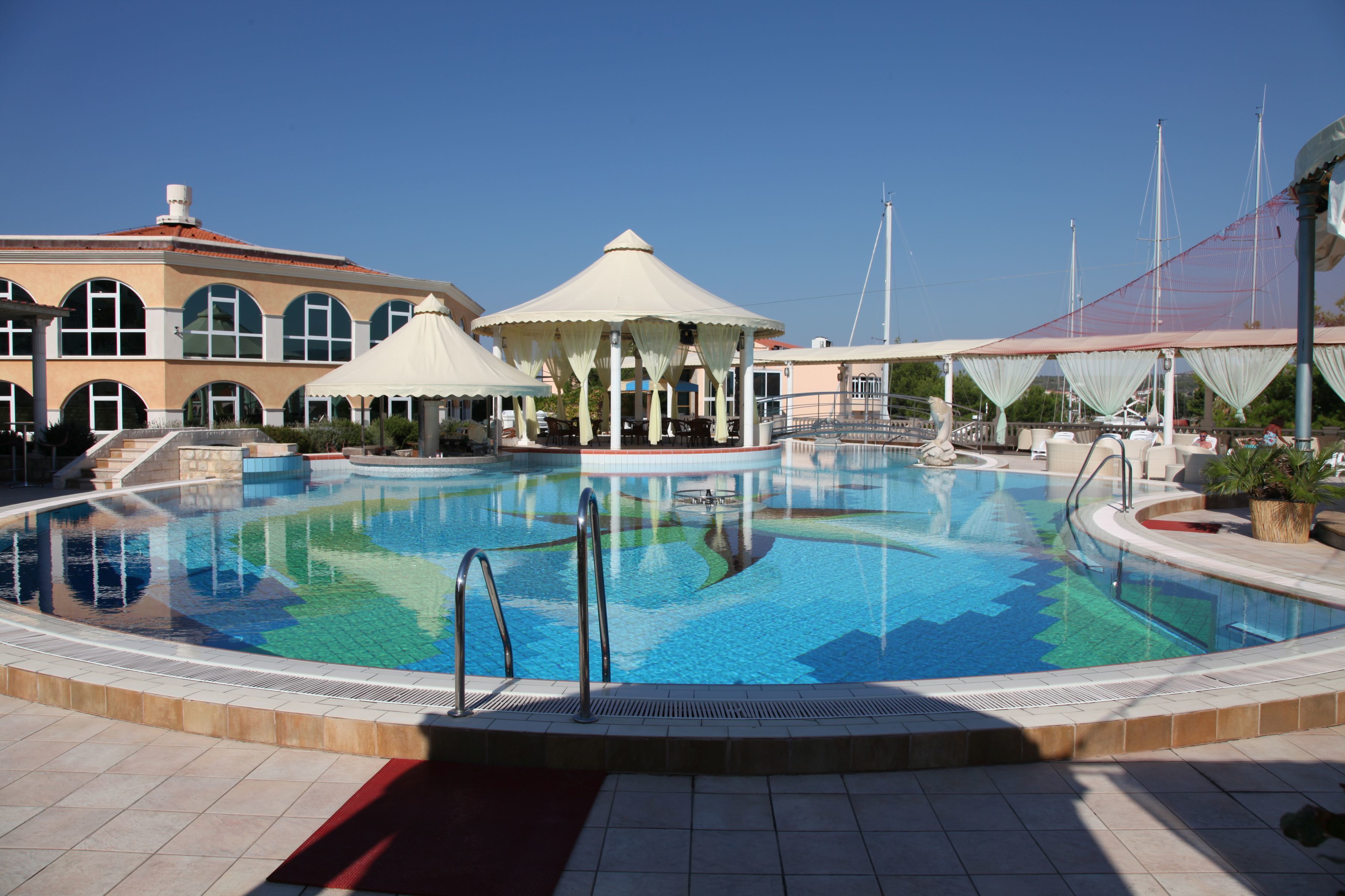 Uma piscina com centro azul translúcido, rodeada por um deck ladrilhado e um edifício com janelas em arco.