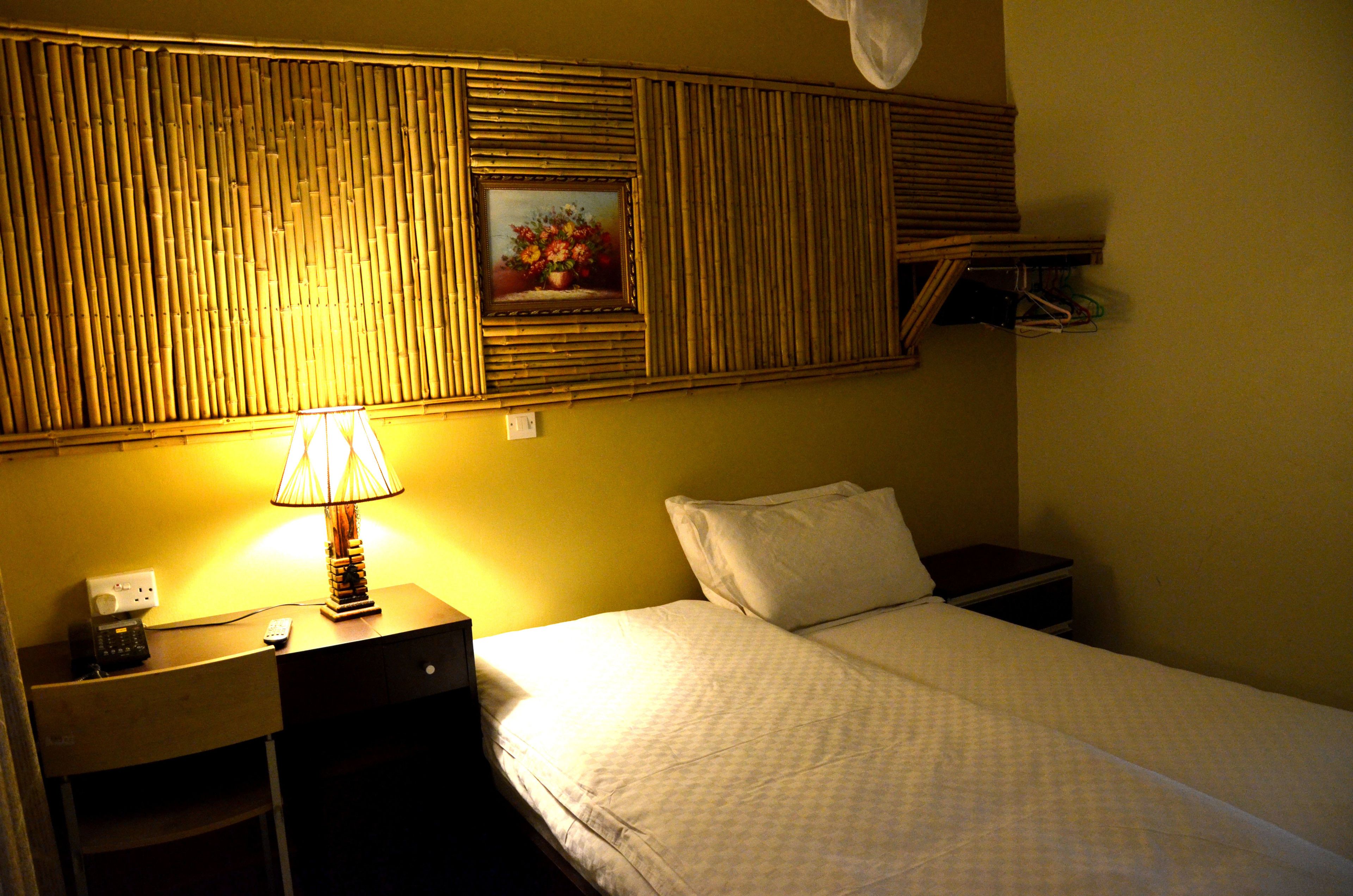 Quarto de hotel com cama, mesa de cabeceira com abajur, pequena escrivaninha com telefone e prateleira na parede com porta-retrato.