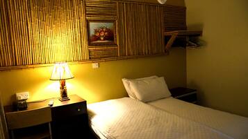 Quarto de hotel com cama, mesa de cabeceira com abajur, pequena escrivaninha com telefone e prateleira na parede com porta-retrato.