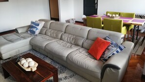 Uma sala de estar com sofá, mesa de centro com objetos decorativos, área de refeições com mesa e cadeiras, e uma porta ao fundo.