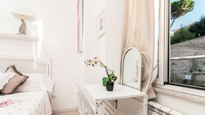 Quarto com cama, mesa de cabeceira com planta em vaso, espelho e janela com vista para uma parede de pedra e árvores.
