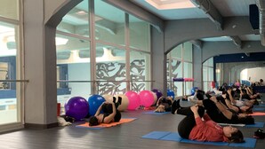 Uma academia espaçosa com amplas janelas, bolas de exercício e pessoas fazendo exercícios no chão.