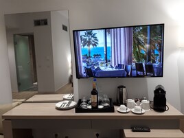 Quarto de hotel com uma televisão de tela grande exibindo uma vista panorâmica, um minibar com copos e uma garrafa, uma cafeteira e um controle remoto na mesa.