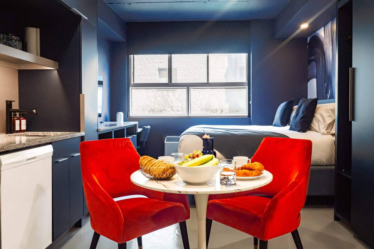 Quarto de hotel compacto com cama, pequeno espaço de cozinha, área de refeições com mesa e cadeiras, e uma grande janela que proporciona luz natural.